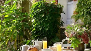 Uma área de refeições ao ar livre com uma mesa posta para o café da manhã, incluindo uma jarra de suco de laranja, uma xícara e uma cesta de doces.