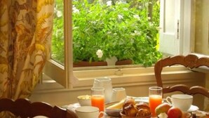 Uma mesa de café da manhã com toalha florida, janela com vista para o verde e diversos itens para o café.