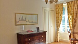 Quarto com cômoda de madeira, quadro com vista urbana emoldurada, lustre, cadeira e janela com cortinas florais.
