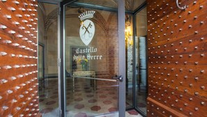 Entrada de um hotel com porta de vidro, parede de madeira com detalhes metálicos e uma placa com a inscrição 'Castello delle Serre'.