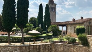 Uma ponte de pedra com arco, um pátio com plantas em vasos e uma torre com relógio ao fundo.