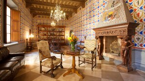 Um quarto ricamente decorado, com teto ornamentado, um candelabro, lareira com uma estátua e uma coleção de estantes de livros.