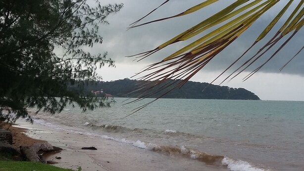 Uma praia com ondas, uma ilha distante e um céu nublado.