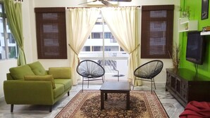 Uma sala de estar com um sofá verde, duas cadeiras pretas, uma mesinha, um tapete estampado e uma TV em uma parede verde.