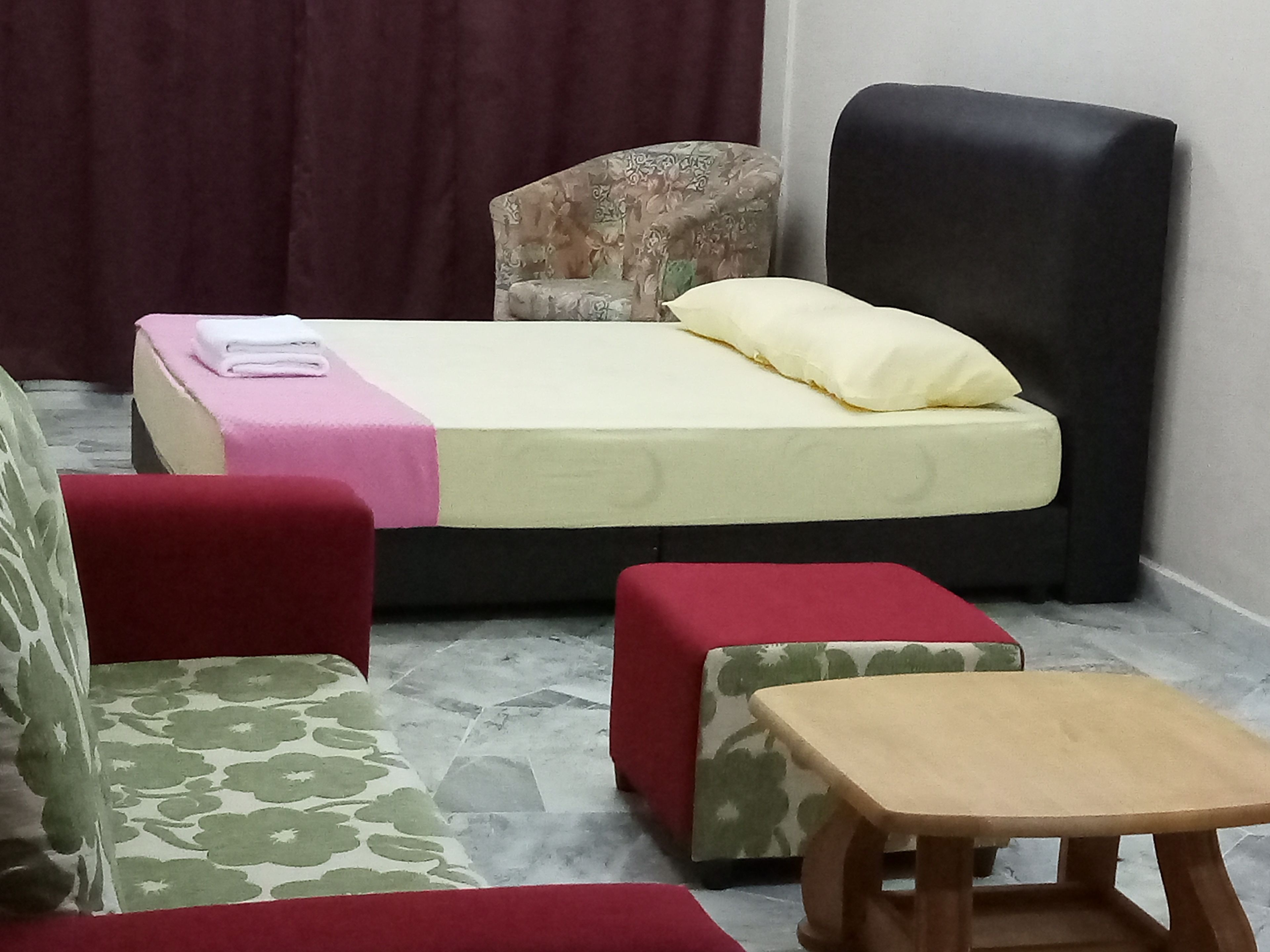 Quarto compacto com uma cama, um sofá vermelho, uma mesinha e uma poltrona estampada.
