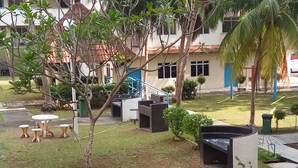 Uma área externa com mesas redondas e bancos, cercada por vegetação e um edifício com telhado em azulejos.