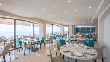 Um restaurante moderno com mesas redondas, cadeiras cor de azul-esverdeado e vista para o mar.