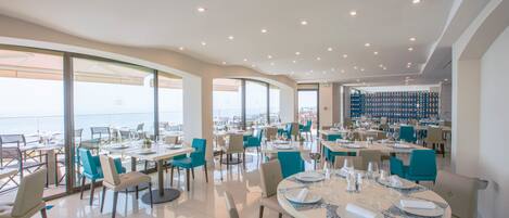 Um restaurante moderno com mesas redondas, cadeiras cor de azul-esverdeado e vista para o mar.
