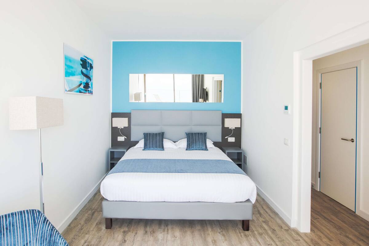 Quarto moderno com uma cama grande, parede azul de destaque, espelho sobre a cama, abajures e uma porta à direita.