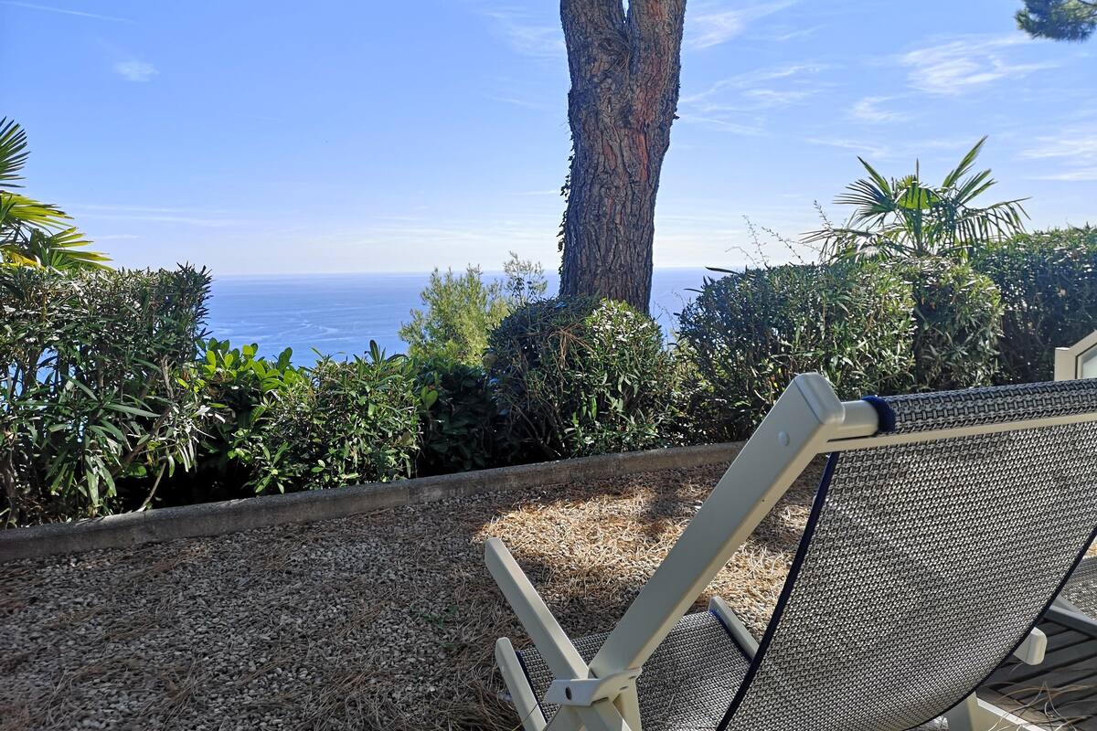 Vista do oceano a partir de um alpendre com cadeira e mesa, cercado por vegetação e uma grande árvore.