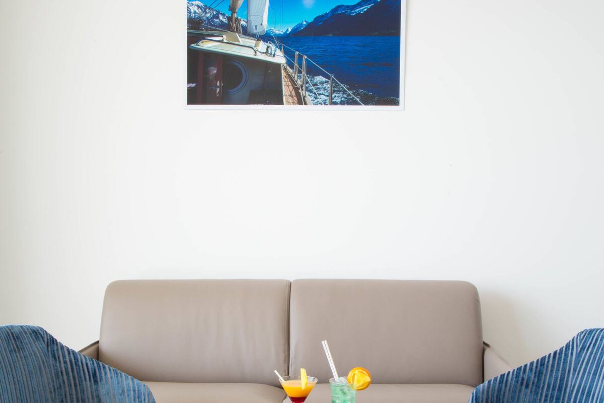 Um quarto com um sofá bege, duas cadeiras azul e branco listradas e uma mesinha redonda com duas bebidas.