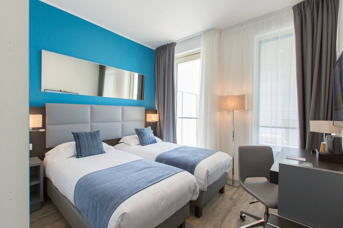 Quarto de hotel com duas camas, uma escrivaninha, uma cadeira e uma janela grande com cortinas.