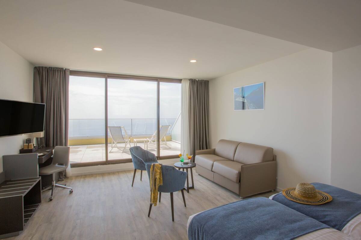 Quarto de hotel com varanda, sofá, televisão, secretária e cadeira.