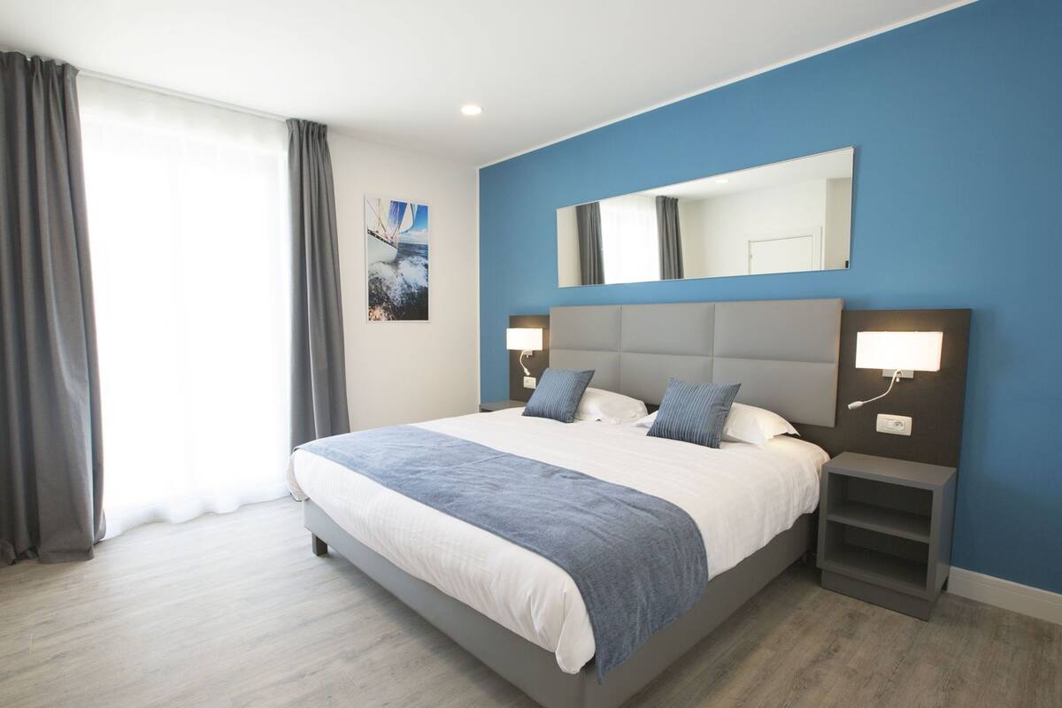 Quarto de hotel moderno com uma cama grande, mesa de cabeceira, espelho e janela com cortinas.