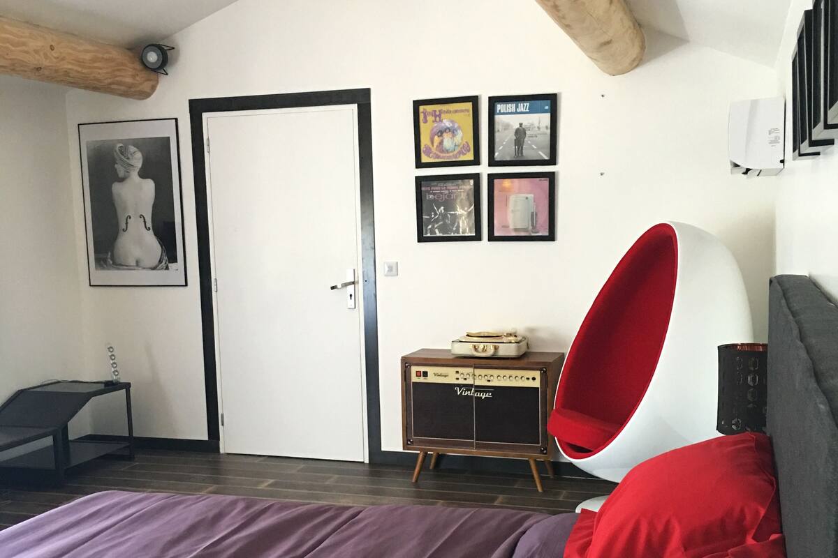 Um quarto com uma cama, uma cadeira vermelha, um amplificador antigo e quadros nas paredes.
