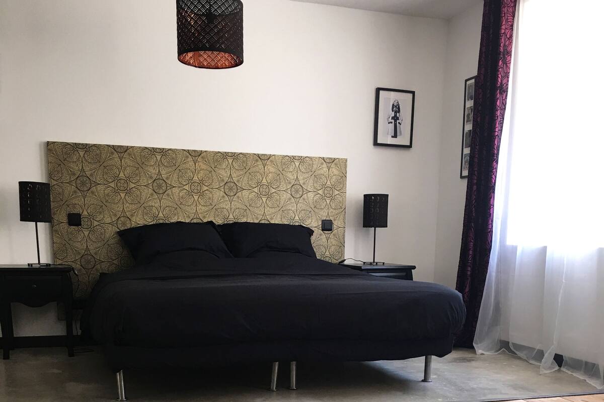 Quarto moderno com uma cama grande, piso de madeira, uma mesa de vidro e cabeceira estampada.