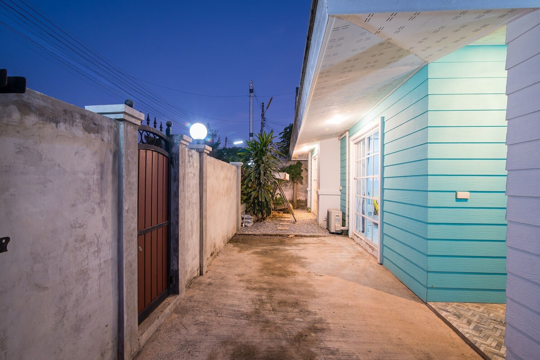 Uma passagem estreita com um caminho de concreto, um portão de metal e um edifício com revestimento azul-turquesa.