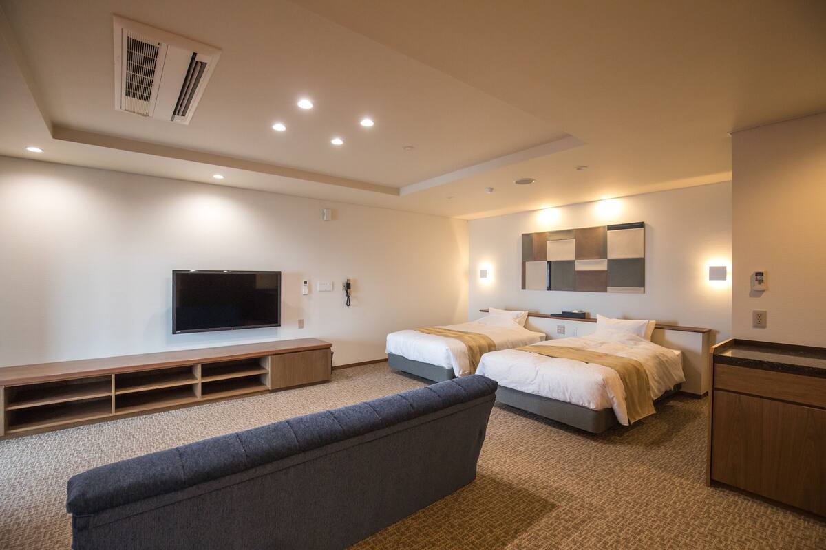 Quarto de hotel com uma grande TV de tela plana, duas camas, um sofá e um armário de madeira.