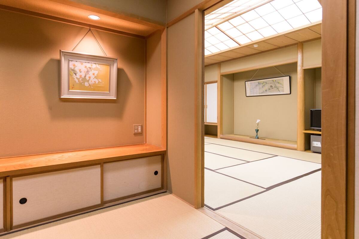 Um quarto japonês tradicional com piso de tatame, um banco de madeira baixo, uma obra de arte emoldurada e uma porta deslizante que dá para outro quarto com um espelho grande e uma televisão.