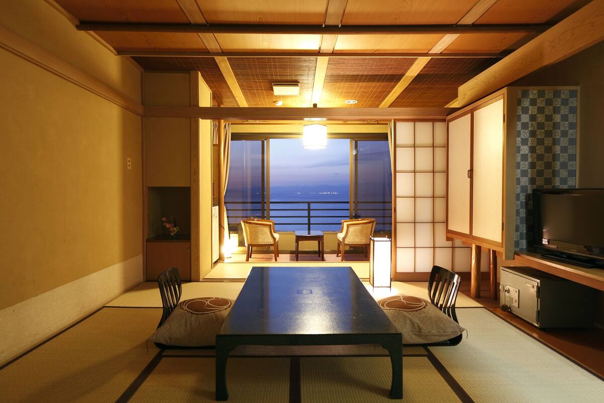 Um quarto japonês tradicional com tatames, uma mesa baixa, cadeiras, uma televisão e vista para o mar.
