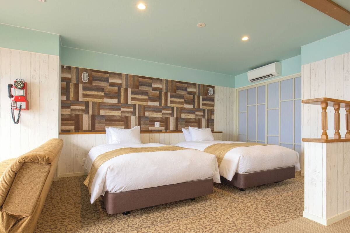 Quarto de hotel com duas camas, cabeceira de madeira, uma pequena escrivaninha e uma cadeira.