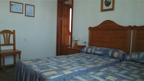Quarto com duas camas, uma cadeira de madeira, uma mesinha de cabeceira com abajur e dois quadros com certificados na parede.