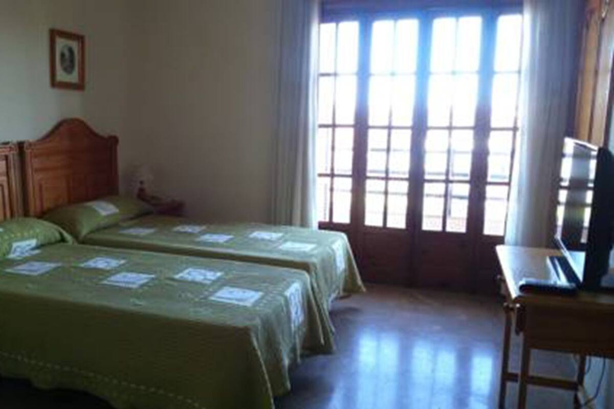 Quarto com duas camas, cabeceira de madeira, uma escrivaninha com um laptop e uma janela com cortinas.