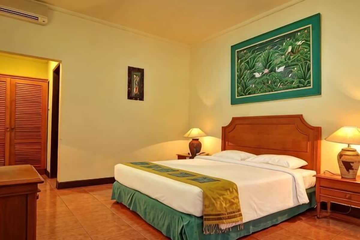 Quarto de hotel com cama, duas luminárias de cabeceira, um quadro na parede e uma porta à esquerda.