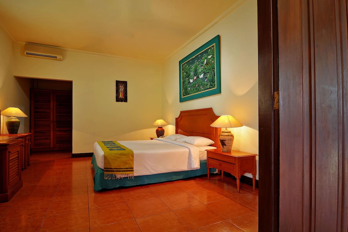 Quarto de hotel com cama, duas mesas de cabeceira com abajures, uma porta de madeira, um ar condicionado fixado na parede e um quadro emoldurado.