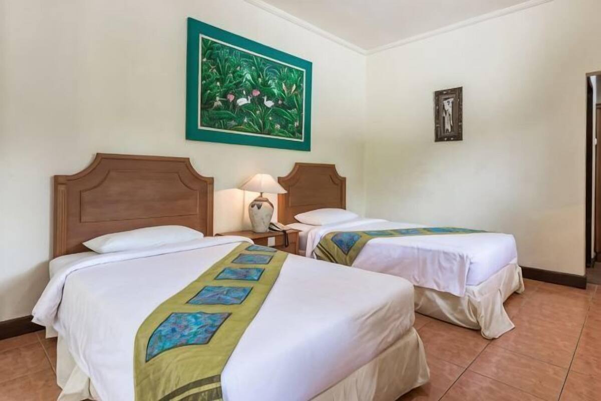 Duas camas de solteiro com cabeceiras de madeira, uma mesa de cabeceira com abajur, um quadro com moldura verde na parede e um quadro acima da porta.