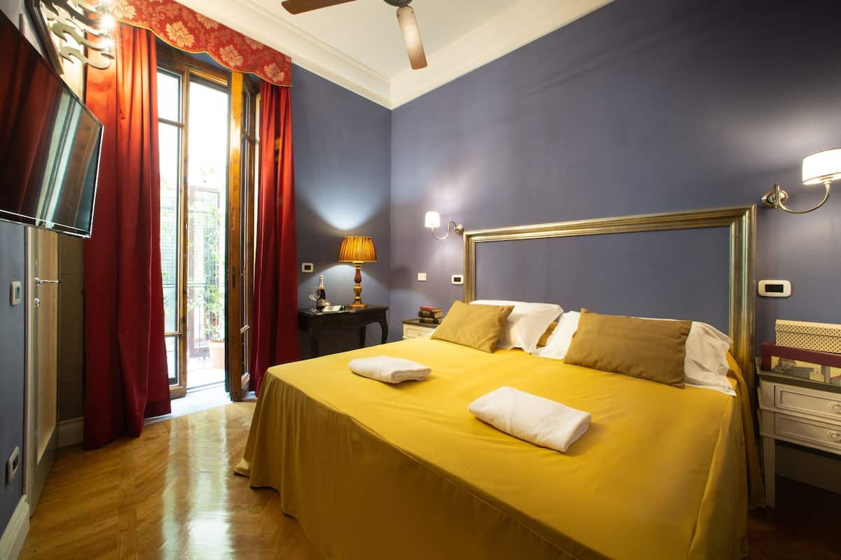 Quarto de hotel com uma cama grande, piso de madeira, ventoinha no teto e uma janela com cortinas vermelhas.