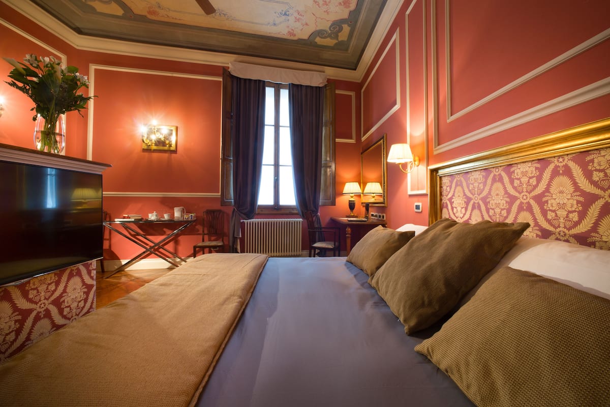 Quarto de hotel com uma cama grande, uma escrivaninha, uma cadeira, uma televisão e uma janela com cortinas.