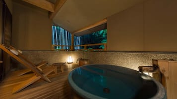 Um quarto de spa com piso de madeira, uma banheira grande, uma cadeira de madeira, uma mesa pequena e uma janela com vista para bambu.