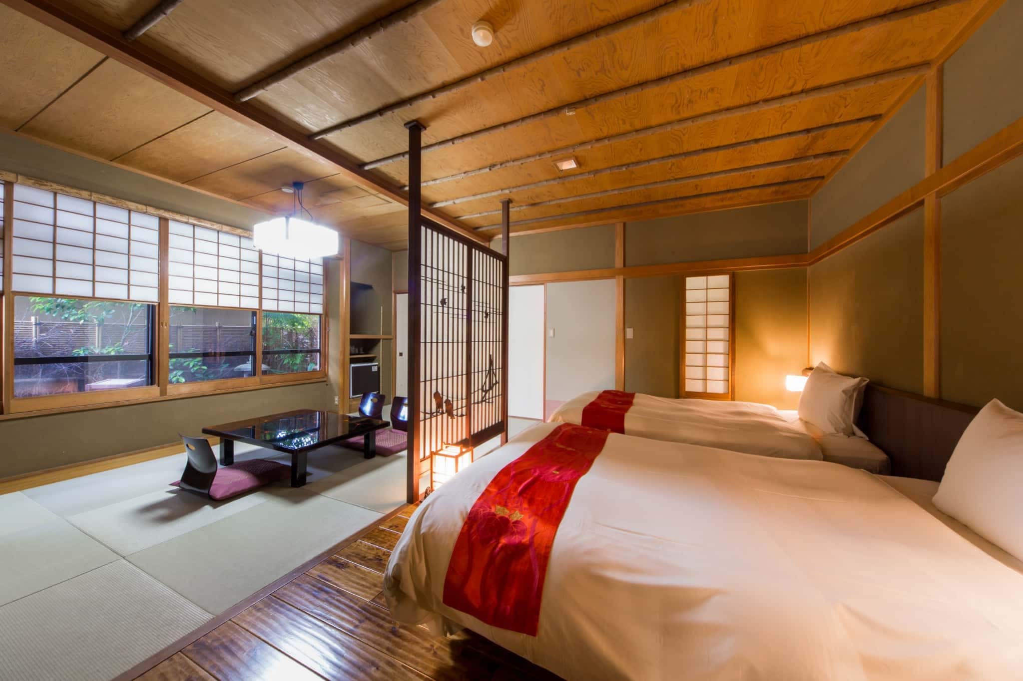 Quarto tradicional japonês com piso de tatame, cama grande com detalhe em vermelho, portas deslizantes e vista para o verde lá fora.
