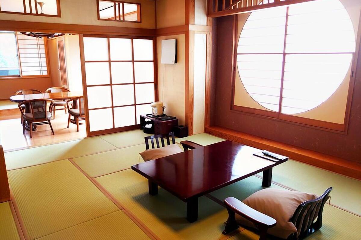 Um espaço com tatames, incluindo uma mesa, um banco e uma divisão tradicional japonesa com portas deslizantes e uma janela grande.