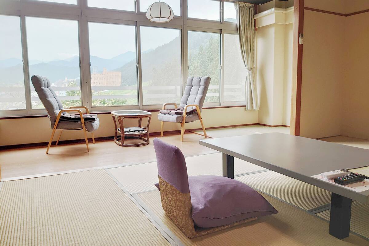Quarto com duas cadeiras, uma mesa pequena e uma janela grande com vista para as montanhas.