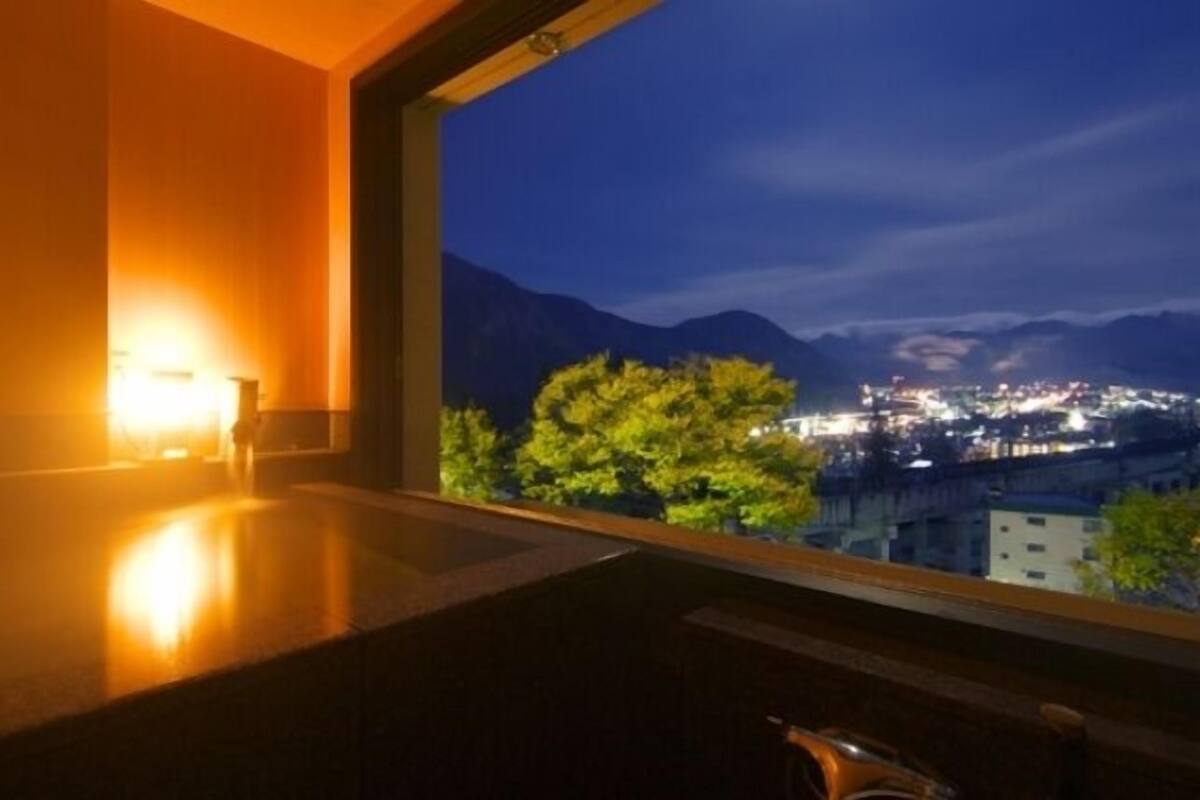 Quarto com uma grande janela com vista para a paisagem urbana à noite, piso de madeira e luminária fixada na parede.