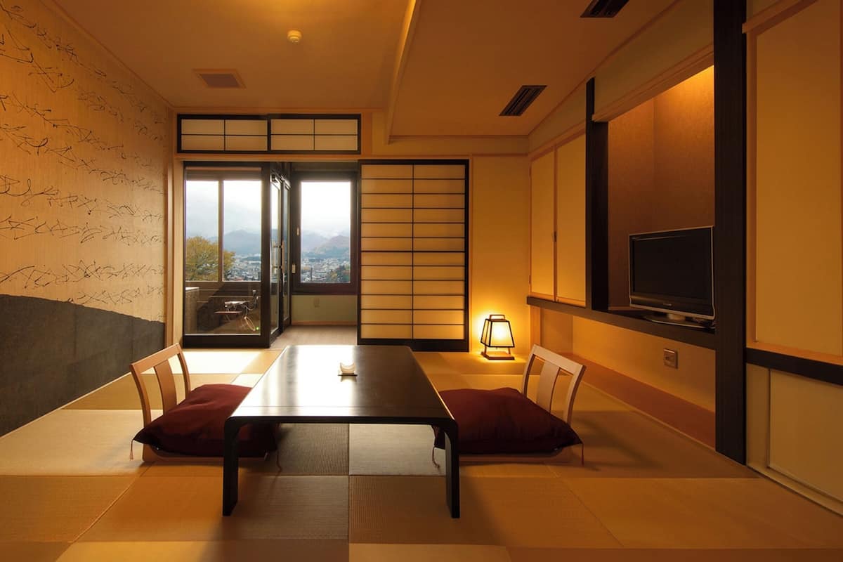 Um quarto japonês tradicional com tatames, uma mesa baixa, portas deslizantes, uma televisão e vista para o horizonte da cidade.
