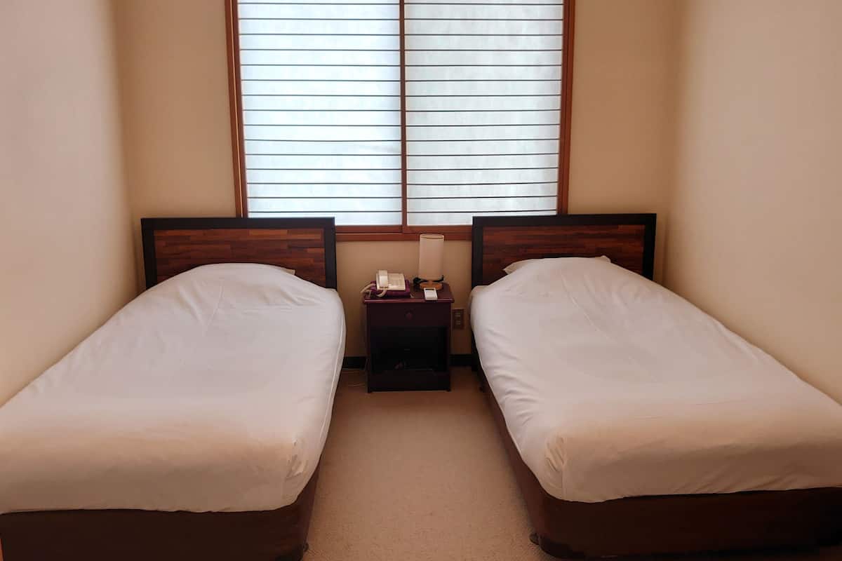 Quarto pequeno e limpo com duas camas de solteiro, mesa de cabeceira com abajur e janela com persianas.