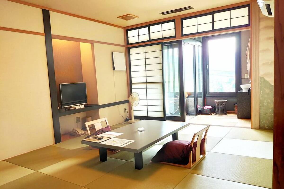 Quarto japonês tradicional com tatames, uma mesa baixa com almofadas, televisão e portas deslizantes que dão para uma varanda.