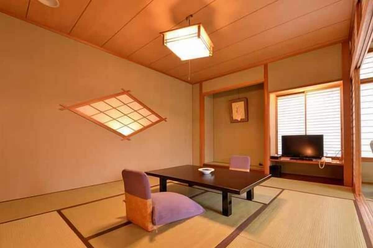 Um quarto japonês tradicional com tatames, uma mesa baixa, um assento de almofada, uma televisão e um painel shoji.