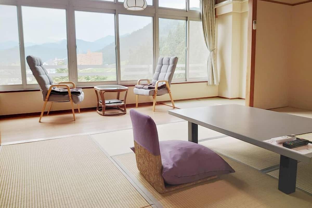 Quarto com esteiras de tatame, uma mesa baixa, duas cadeiras e vista para montanhas através de amplas janelas.