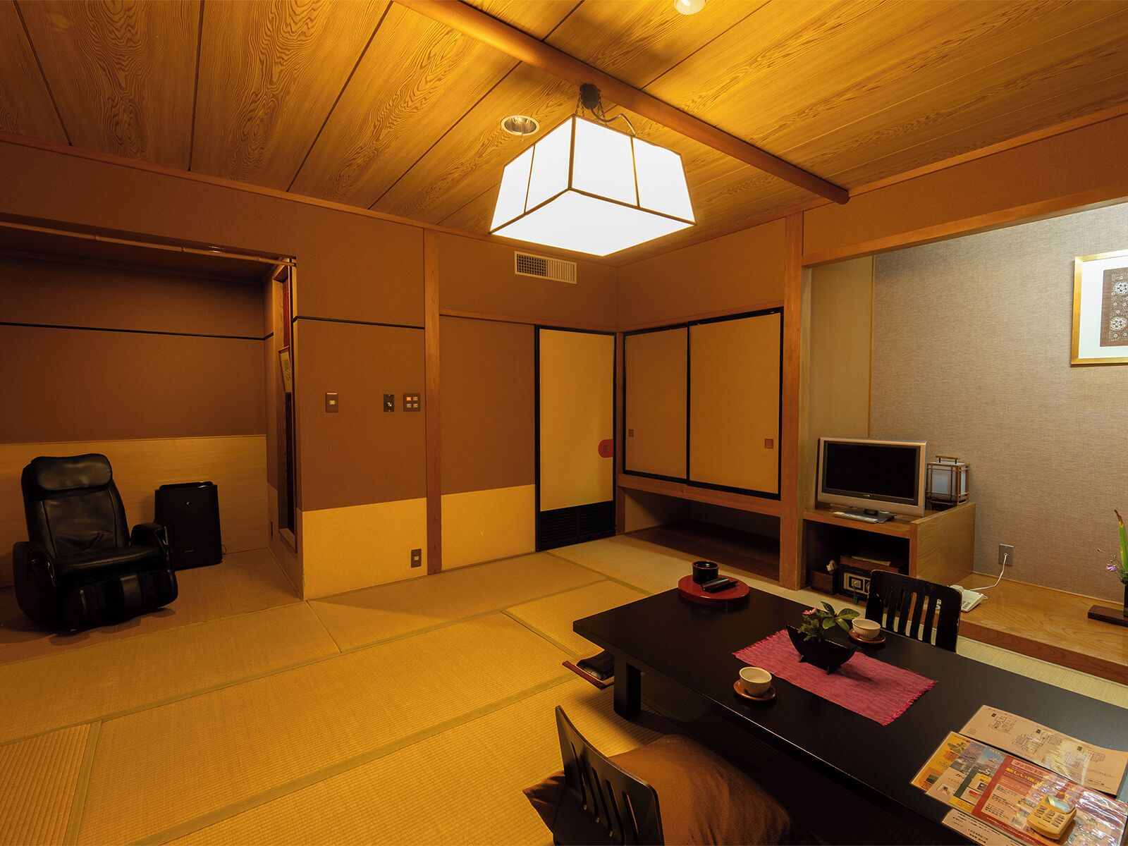 Quarto tradicional japonês com piso de tatame, uma mesa baixa com jogo americano vermelho, uma televisão sobre suporte de madeira e um sistema de som.