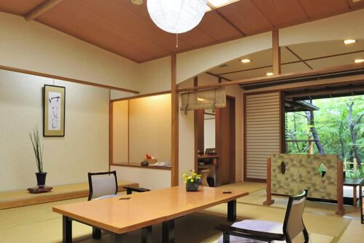 Um quarto em estilo japonês tradicional com tatames, uma mesa de madeira, cadeiras, um biombos shoji e vista para o verde lá fora.