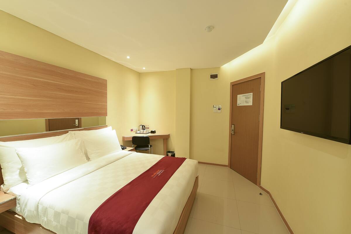 Quarto de hotel com cama, escrivaninha, cadeira, televisão e porta.
