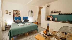 Quarto com cama, mesa de cabeceira e uma pequena escrivaninha com cadeira. Há também uma cozinha compacta com pia, fogão e prateleiras.