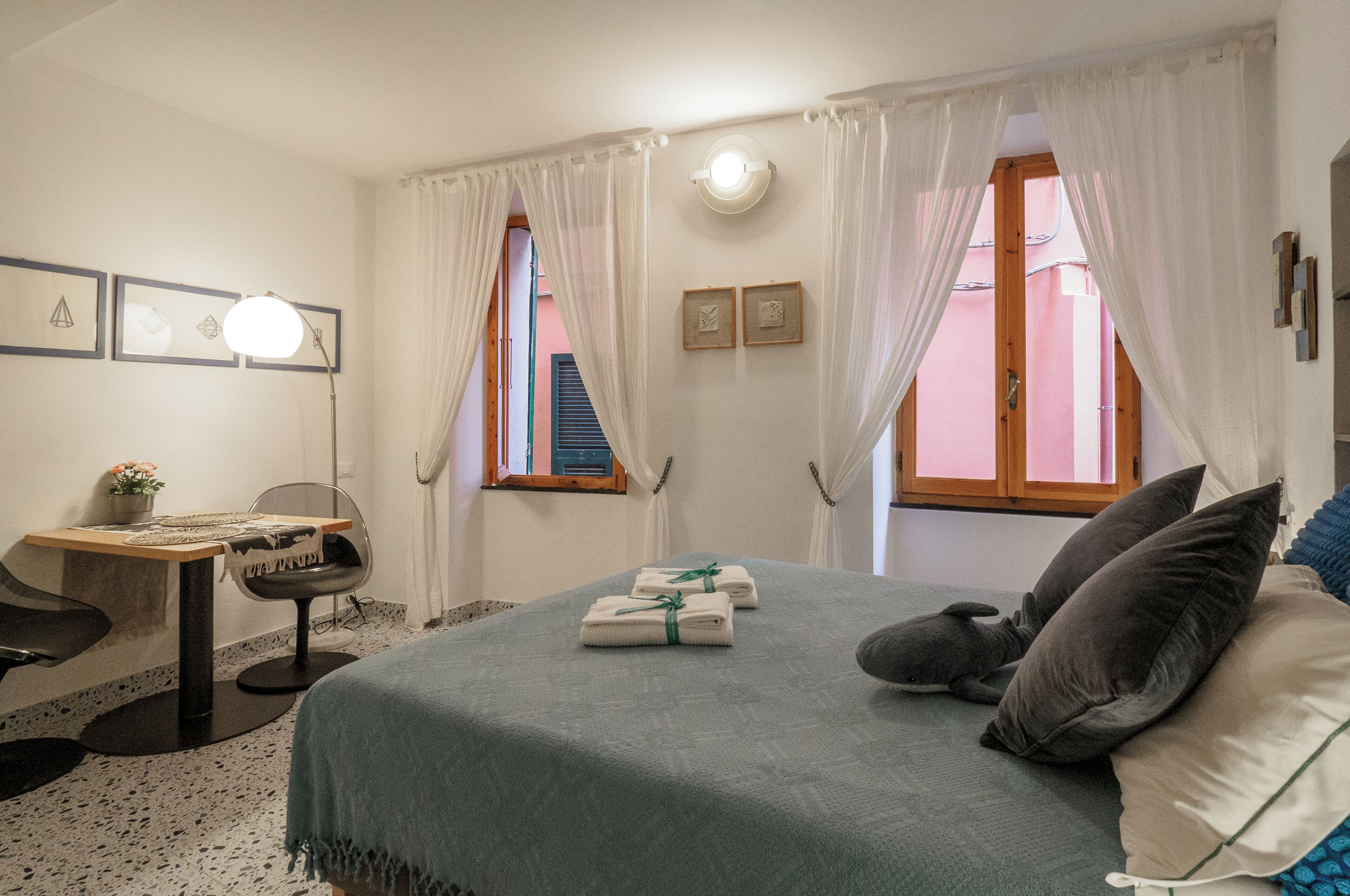 Quarto com cama, almofadas, escrivaninha com cadeira, luminária e cortinas.