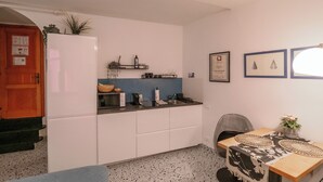 Uma cozinha compacta com armários brancos, micro-ondas e pia. Há uma porta de madeira e uma pequena mesa com uma planta.