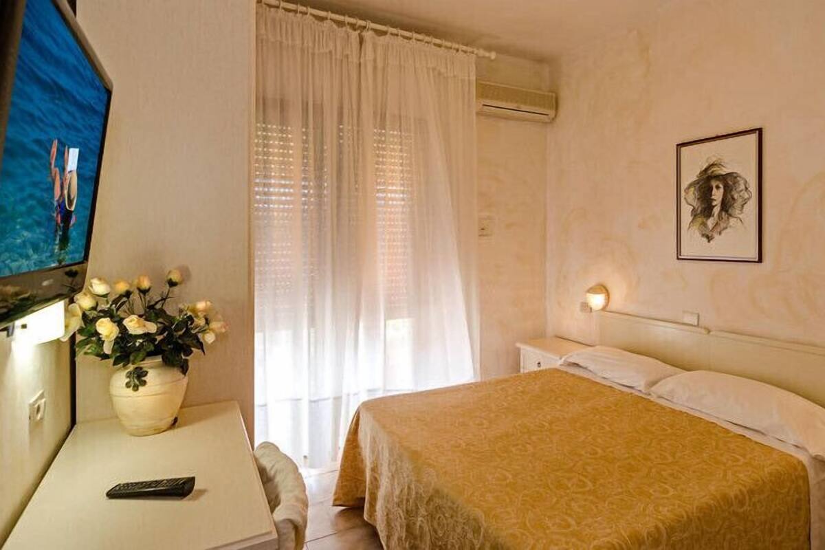 Quarto de hotel com cama, escrivaninha com controle remoto, planta em vaso, televisão na parede e quadro decorativo.