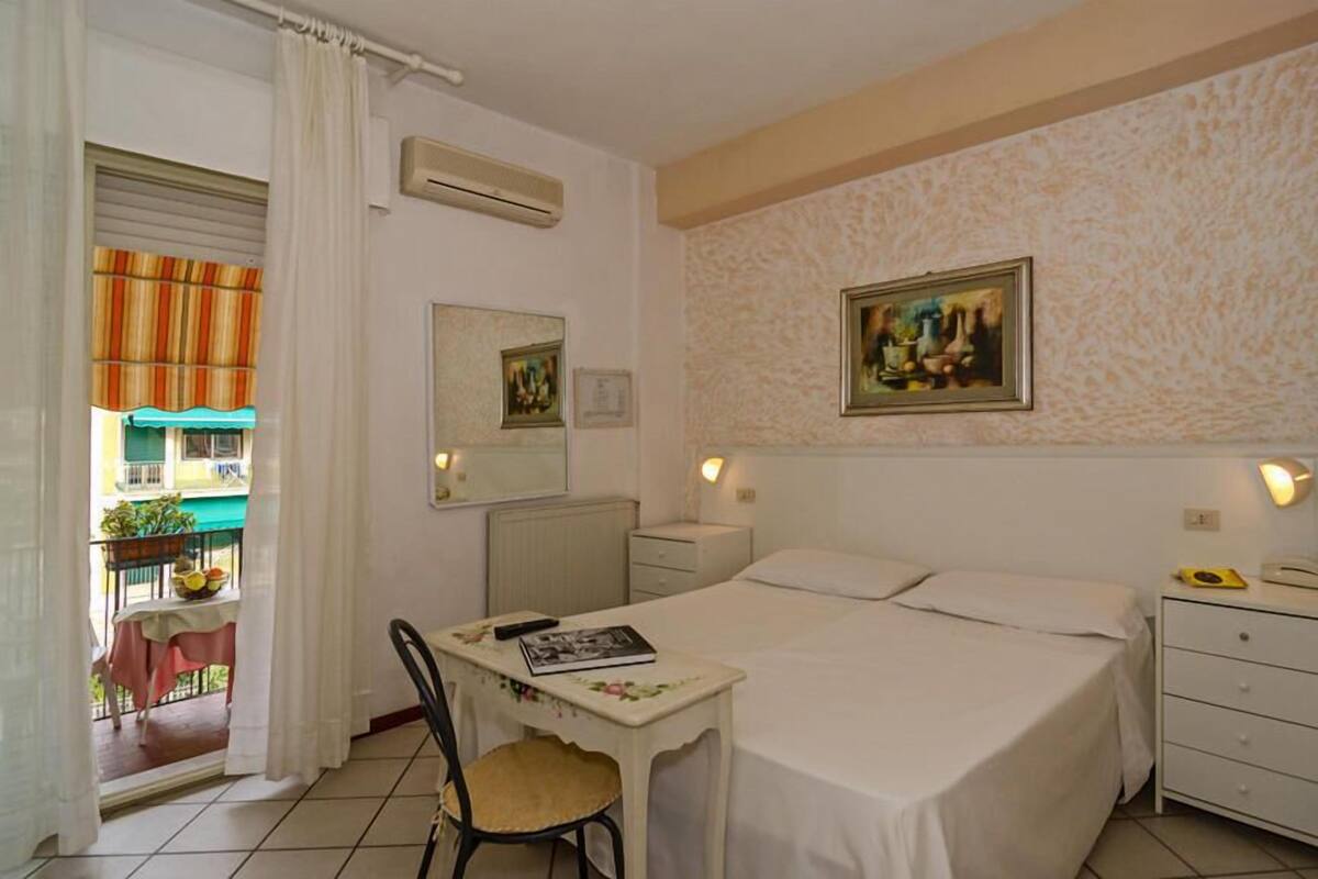 Quarto de hotel com cama, mesas de cabeceira, escrivaninha com cadeira, janela com cortinas e varanda com mesa e cadeiras.